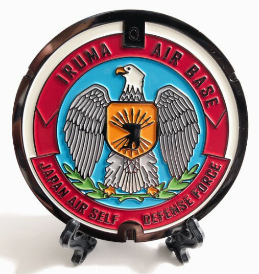 「入間基地」のエンブレムをモチーフとした デザイン。 盾中の矢は部隊数、文字は エンブレムのベースとなったジョンソン基地 の「J」と言われています。 サイズ：径90×厚5mm 材　質：亜鉛合金 スタンド＆化粧箱付き