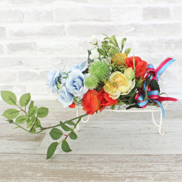 ソファーの形の白いかごに入った、青やオレンジのカラフルなバラの花束の風水アレンジです。サイズ：約高さ15×幅25×奥行11cm＊商品の仕様は変更になる場合がございます＊受注制作のため数日お時間をいただく場合がございます シルクフラワーとは、...